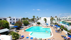 Área de piscina do resort com vários espreguiçadeiras e guarda-sóis, cercada por edifícios brancos e palmeiras.