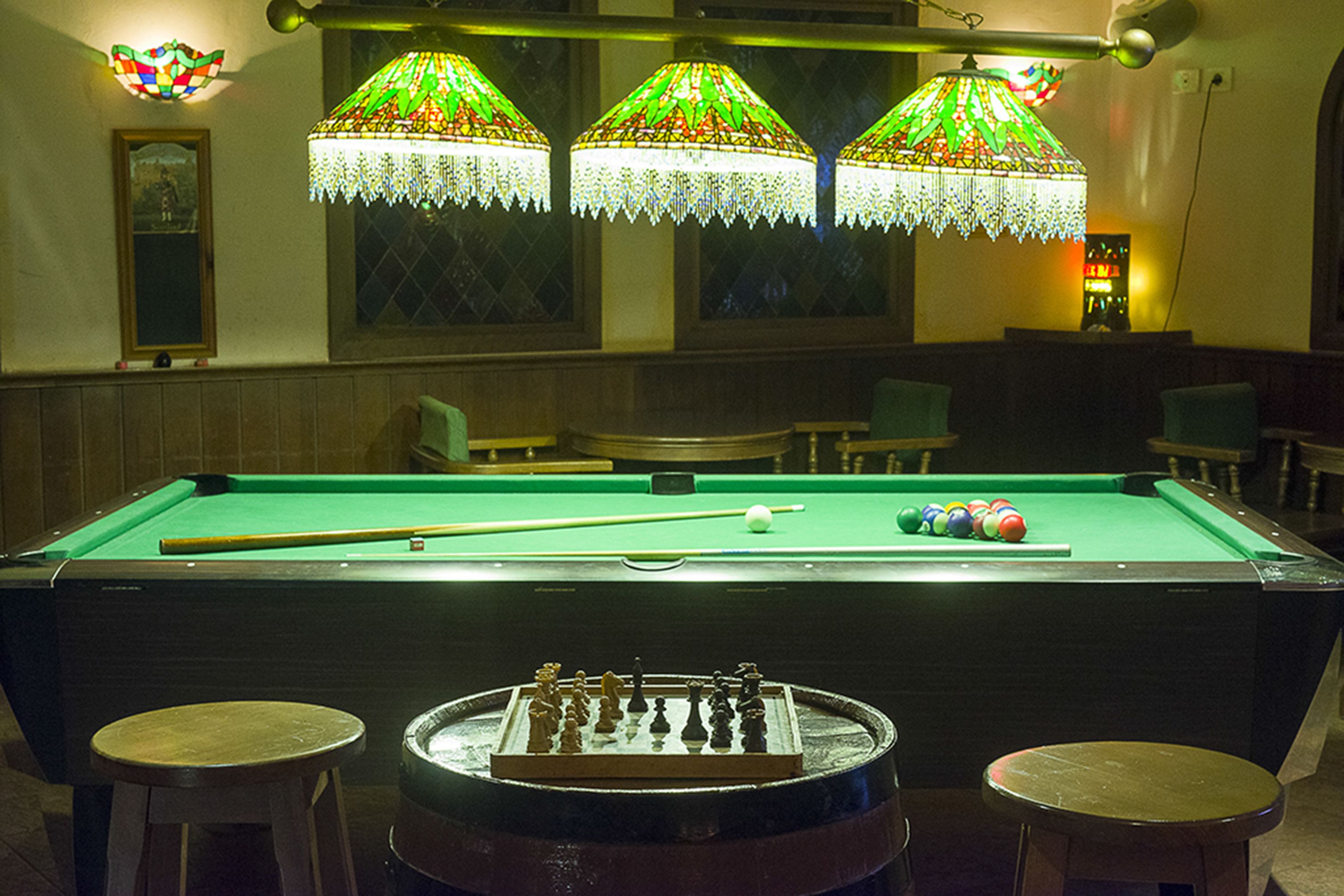 Uma mesa de bilhar com um jogo de xadrez no centro, rodeada por banquetas, sob três luminárias pendentes.