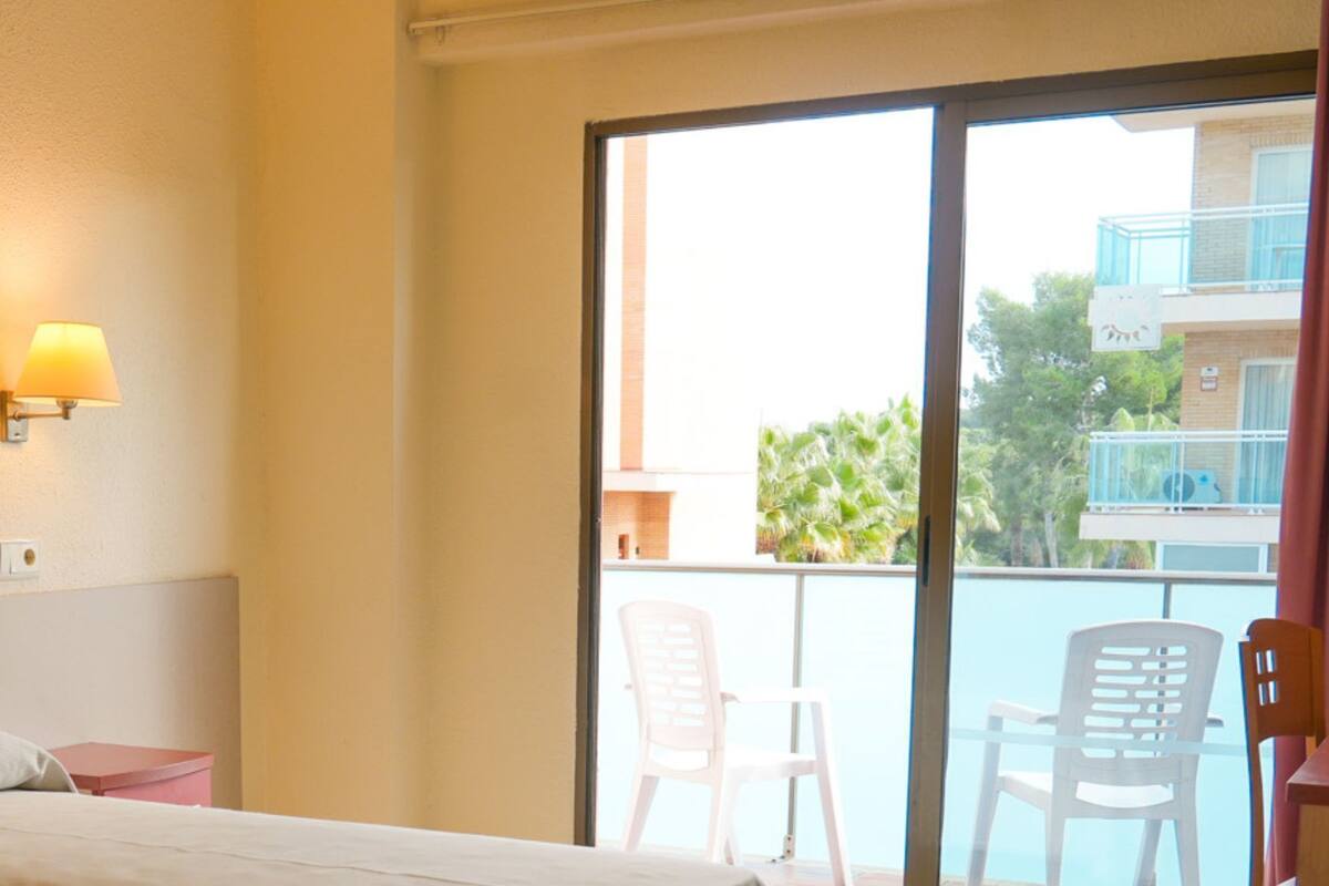 Quarto de hotel com cama, mesa de cabeceira, abajur e vista para uma varanda com cadeiras e um prédio ao fundo.