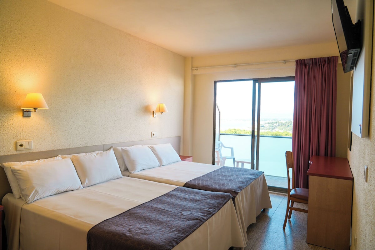 Quarto de hotel com duas camas, uma escrivaninha e vista para uma varanda.