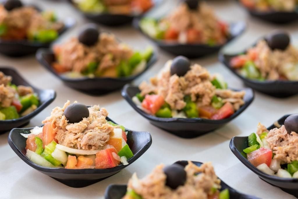 Vários recipientes pretos contêm uma salada de atum, legumes e azeitona preta.