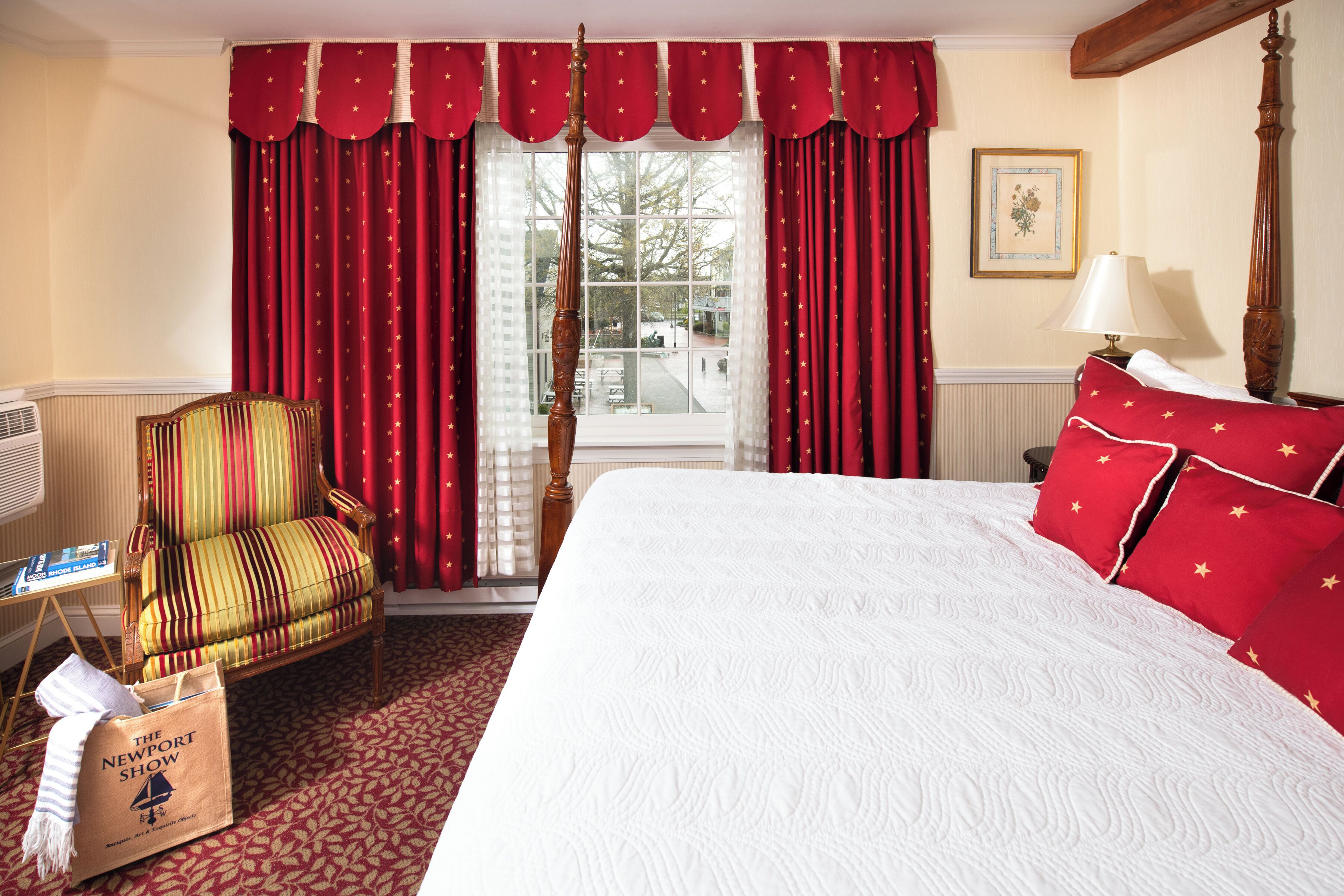 Quarto de hotel com uma cama grande, cortinas vermelhas, uma poltrona listrada e vista para o exterior.