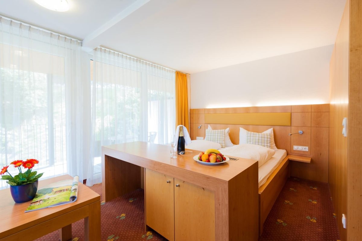 Quarto de hotel com cama, mesa de cabeceira e escrivaninha com cadeira. Há uma janela com cortinas, uma planta em vaso e uma fruteira na escrivaninha.