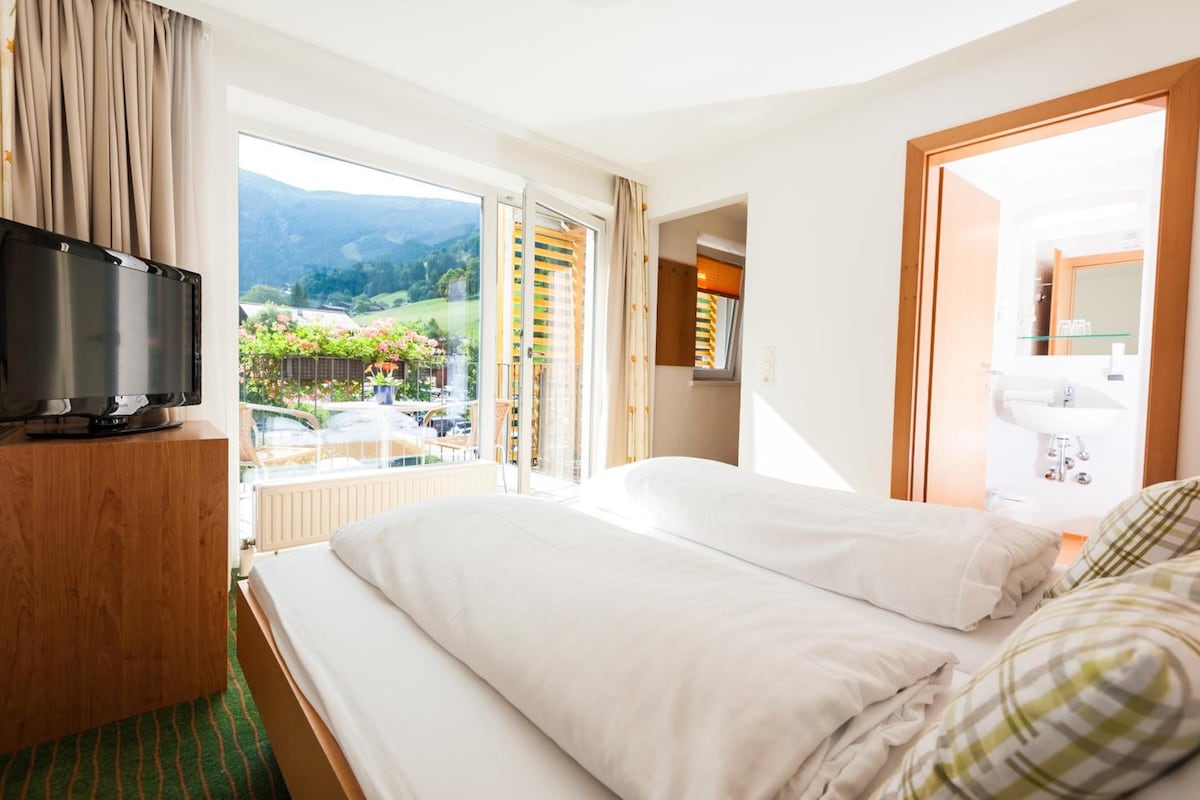 Quarto de hotel com uma cama, televisão sobre um suporte de madeira e vista para uma paisagem montanhosa pela janela.