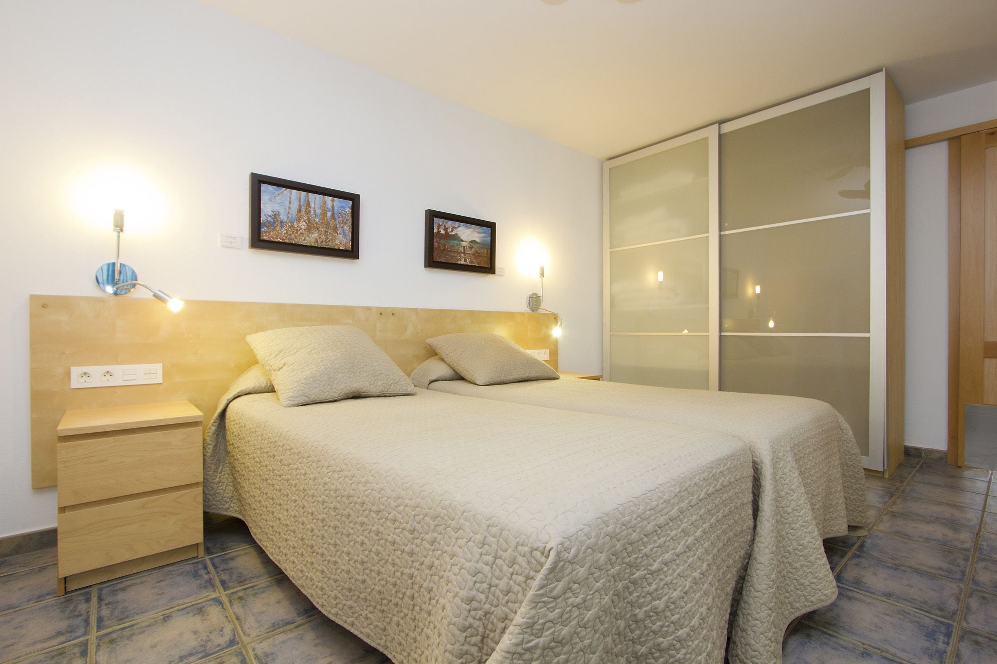 Quarto com cama, mesas de cabeceira, luminárias fixadas na parede e um armário com porta de vidro deslizante.