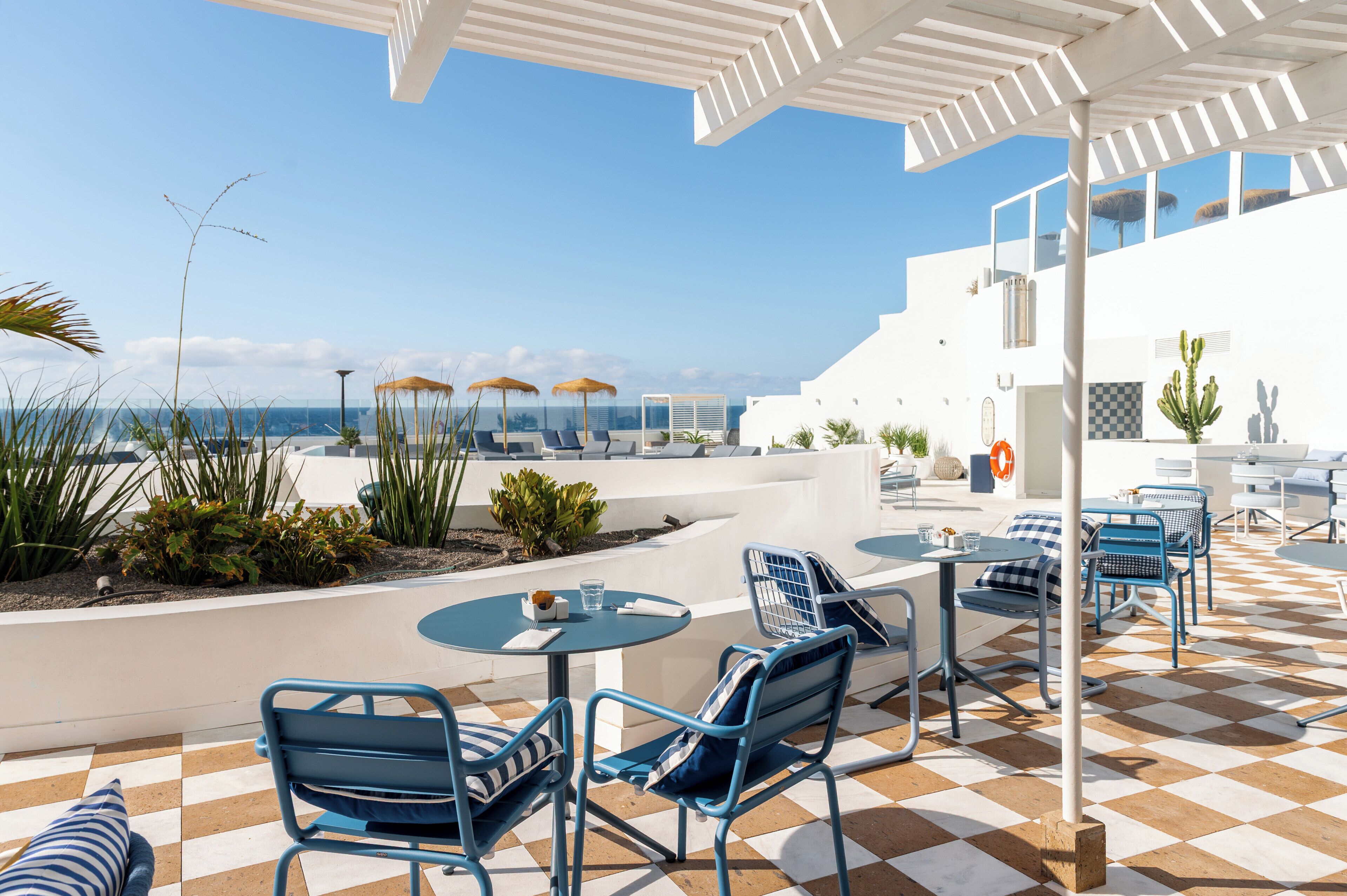 Um terraço com mesas ao ar livre, vista para o mar e um edifício branco com design moderno.