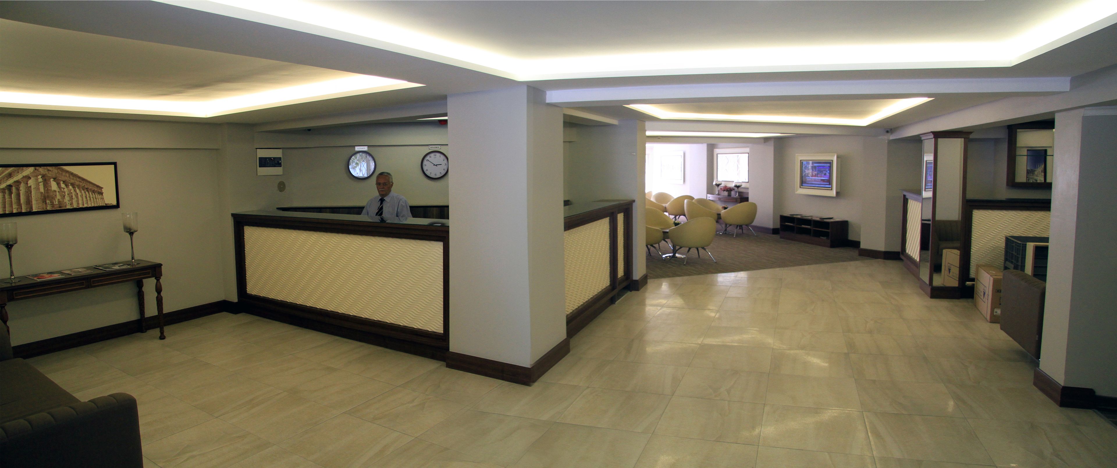 Uma área de recepção de hotel, com balcão de atendimento, cadeiras, relógios e uma grande moldura na parede.