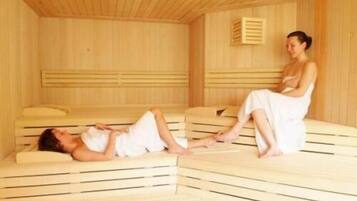Sala de sauna com bancos de madeira e assentos, uma pessoa deitada e outra sentada de pernas cruzadas.