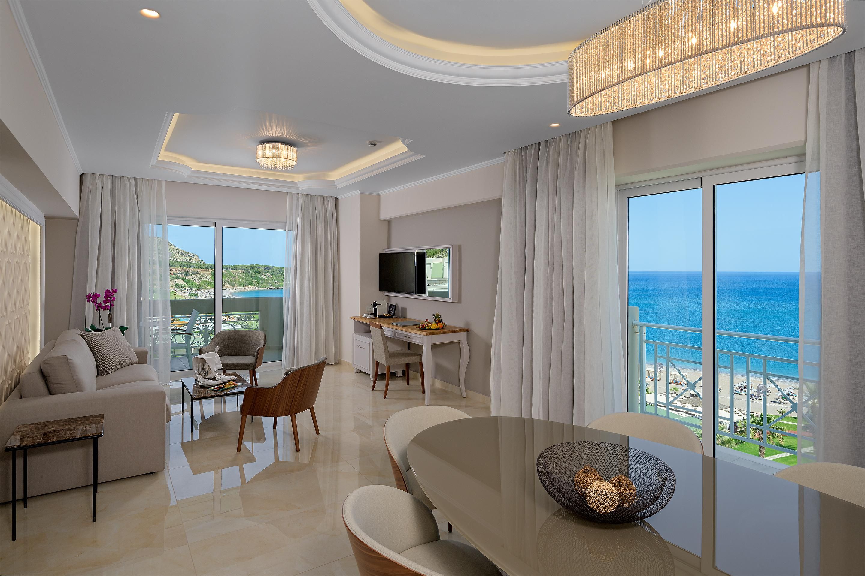 Uma sala de estar moderna com uma grande mesa de jantar, um sofá e uma televisão. O quarto possui janelas amplas com vista para o oceano.
