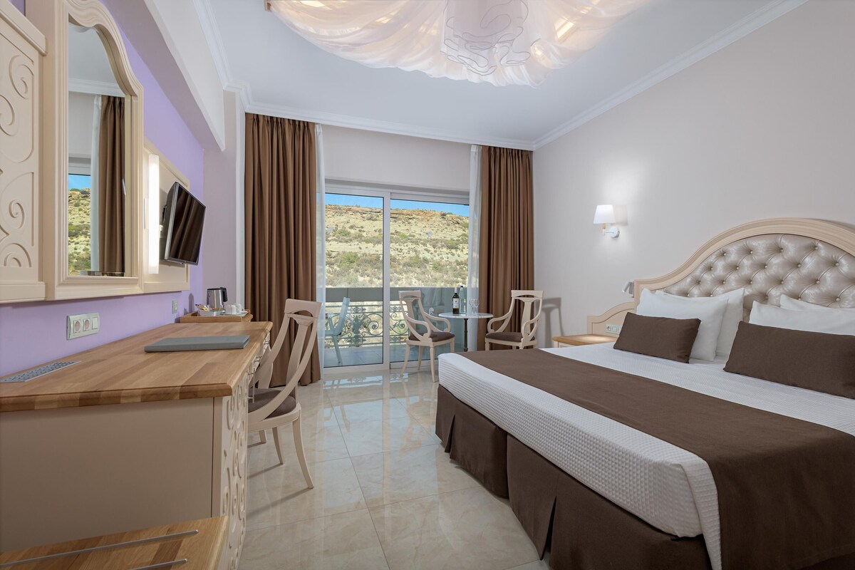 Quarto de hotel com uma cama, escrivaninha, cadeira, televisão e varanda com vista para as colinas.