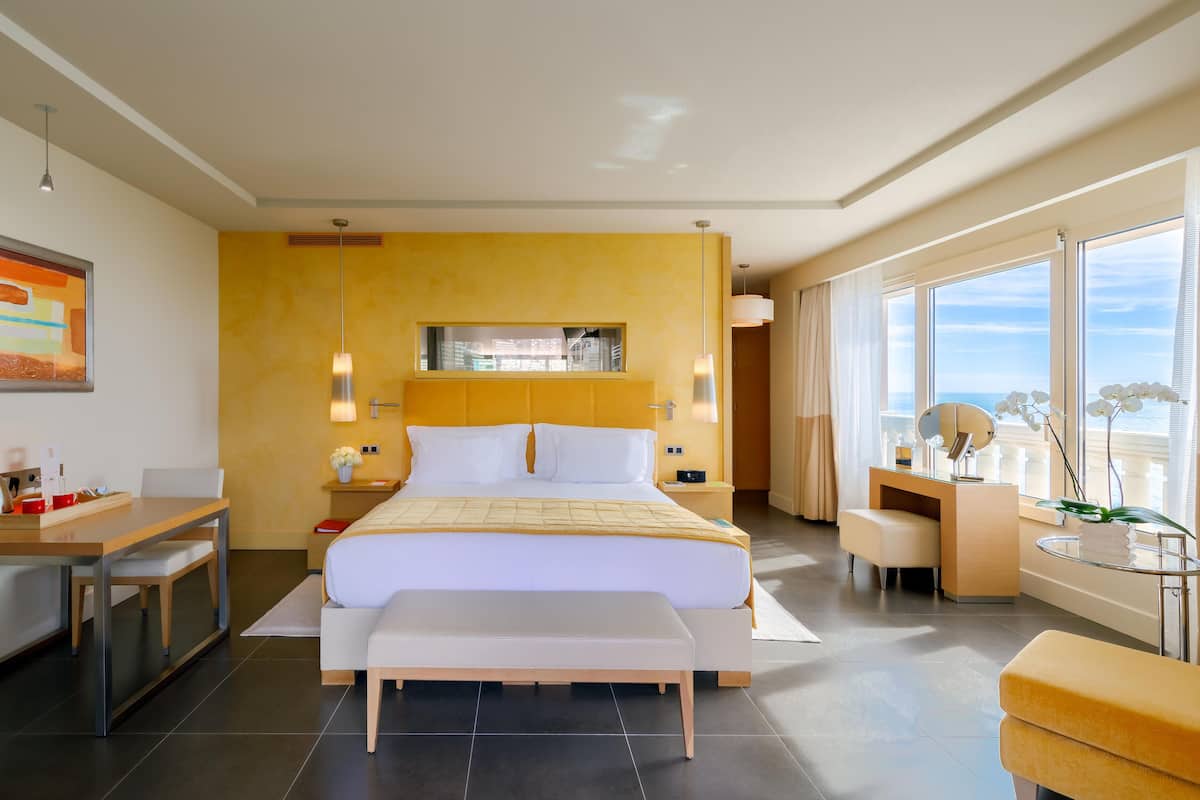 Quarto de hotel moderno com uma cama grande, mesas de cabeceira, uma escrivaninha e vista para a cidade.