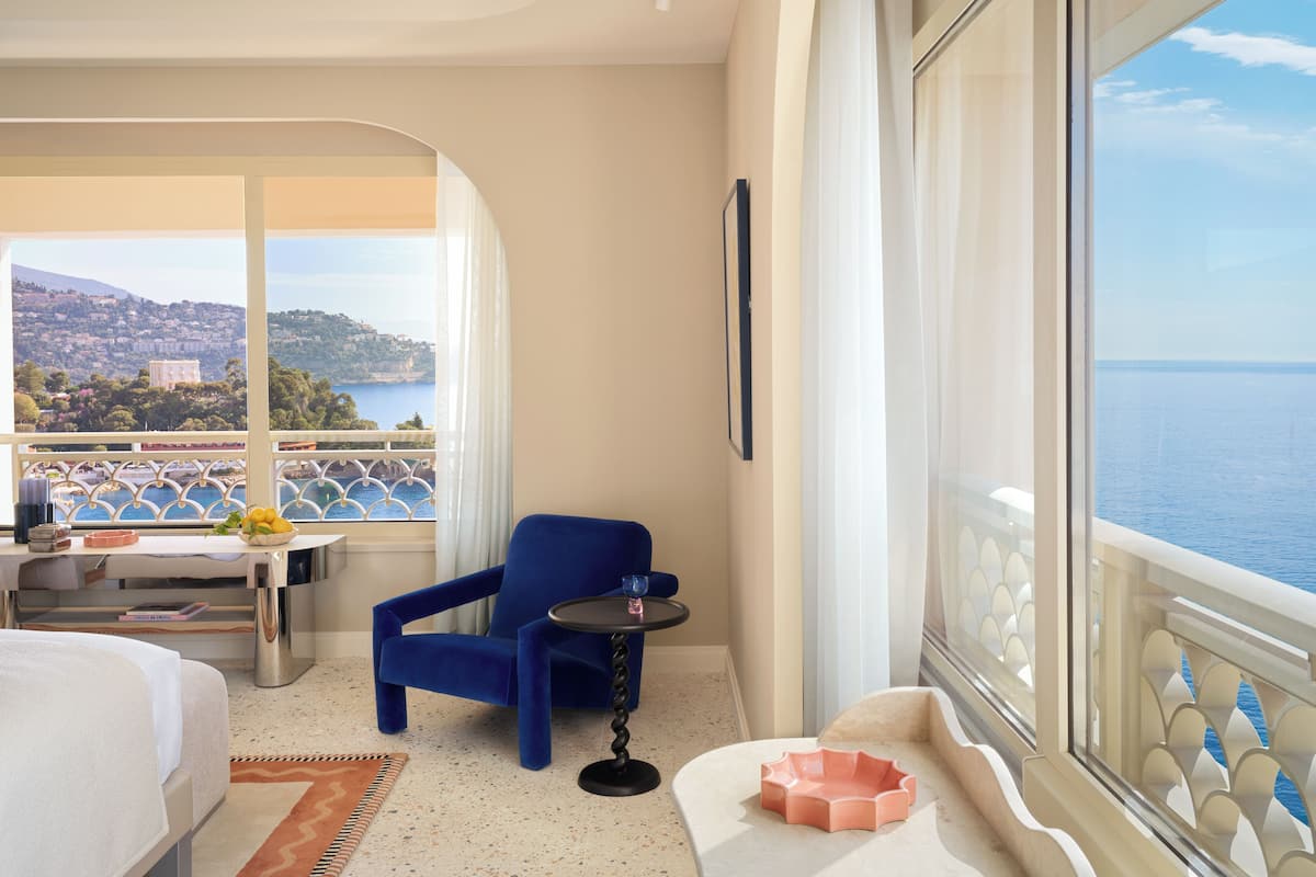 Varanda com vista para o mar e para a cidade. Há uma cadeira azul, uma mesinha com um suporte rosa e uma cama com cabeceira branca.