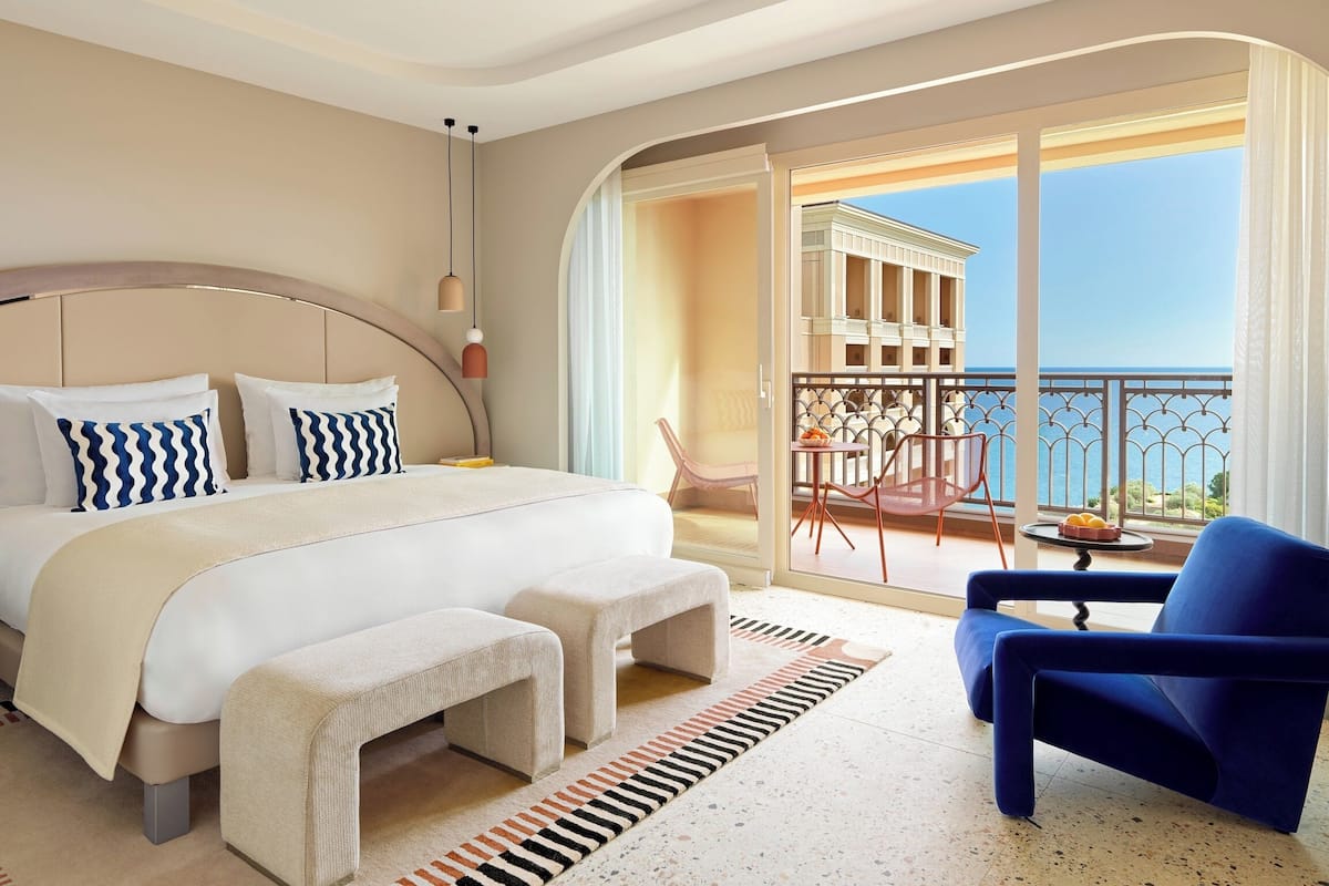 Quarto de hotel moderno com uma cama grande, varanda com vista para o mar e área de refeições com mesa e cadeiras.