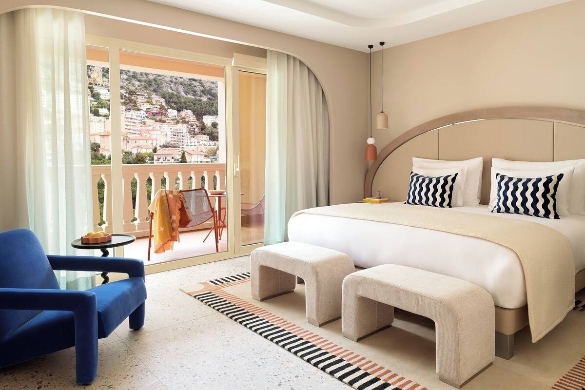 Quarto de hotel com uma cama grande, uma poltrona azul, uma mesinha e vista para uma encosta através de uma varanda.