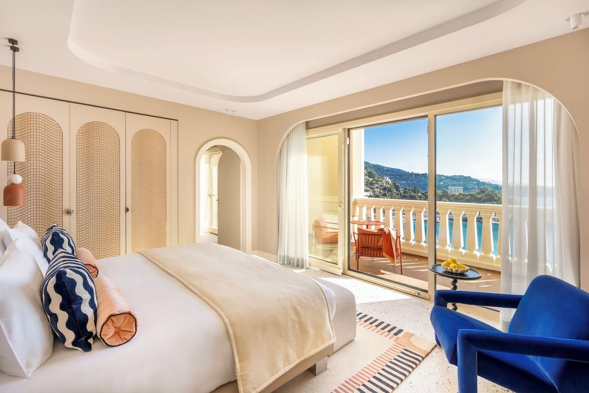 Quarto de hotel com uma cama, uma cadeira azul, uma varanda com mesa e cadeiras, e vista para a paisagem.