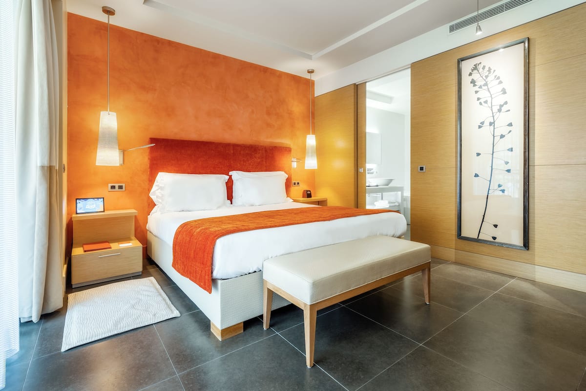 Quarto de hotel moderno com uma cama grande, uma mesinha de cabeceira, um banquinho e uma obra de arte na parede.