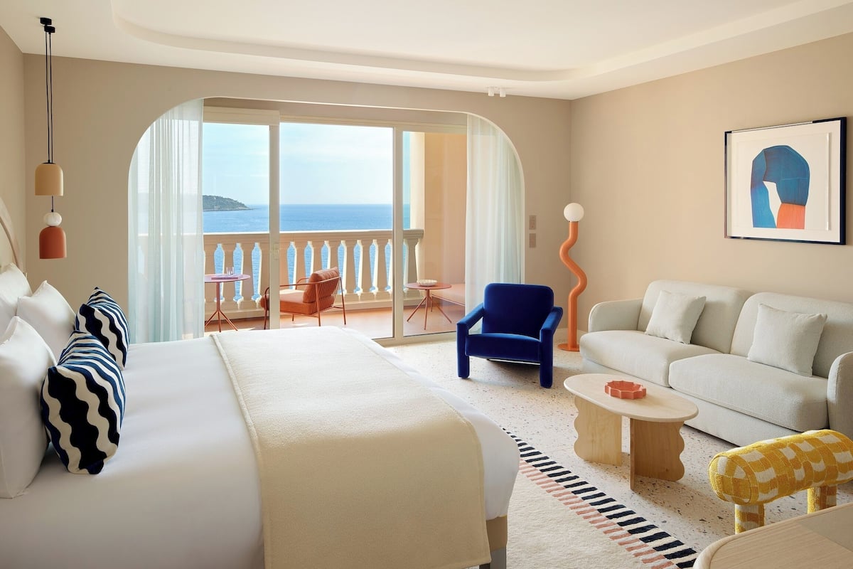 Quarto de hotel com uma cama grande, um sofá, uma poltrona azul, uma mesa pequena e vista para o mar através de uma varanda.