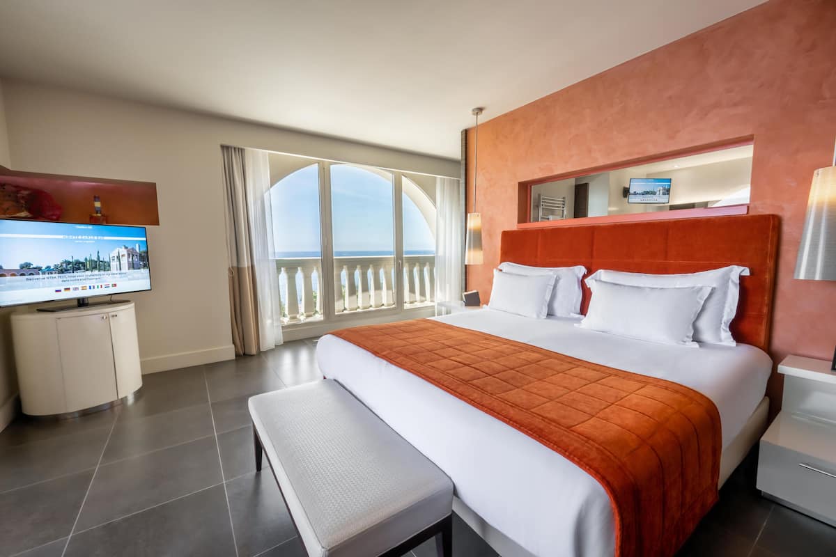 Quarto de hotel com uma cama grande, televisão, um banco e varanda com vista.