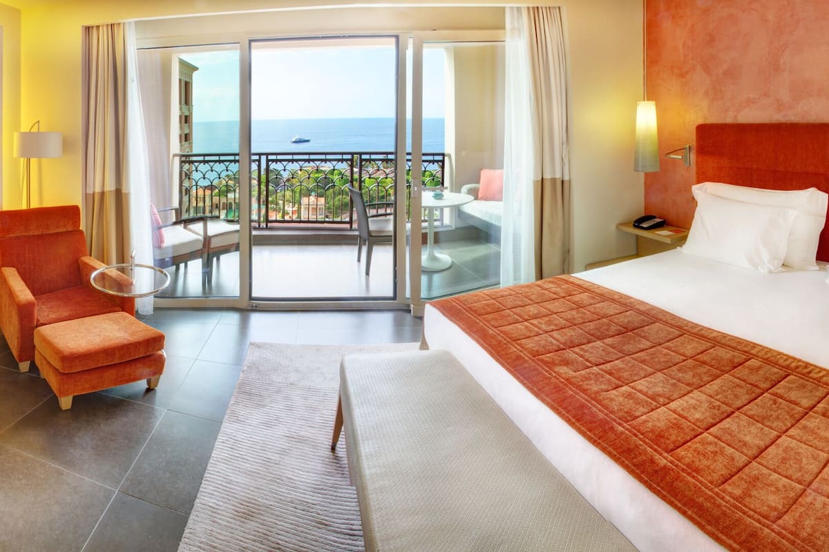 Quarto de hotel com uma cama grande, uma cadeira, uma mesinha e uma varanda vista para o mar.