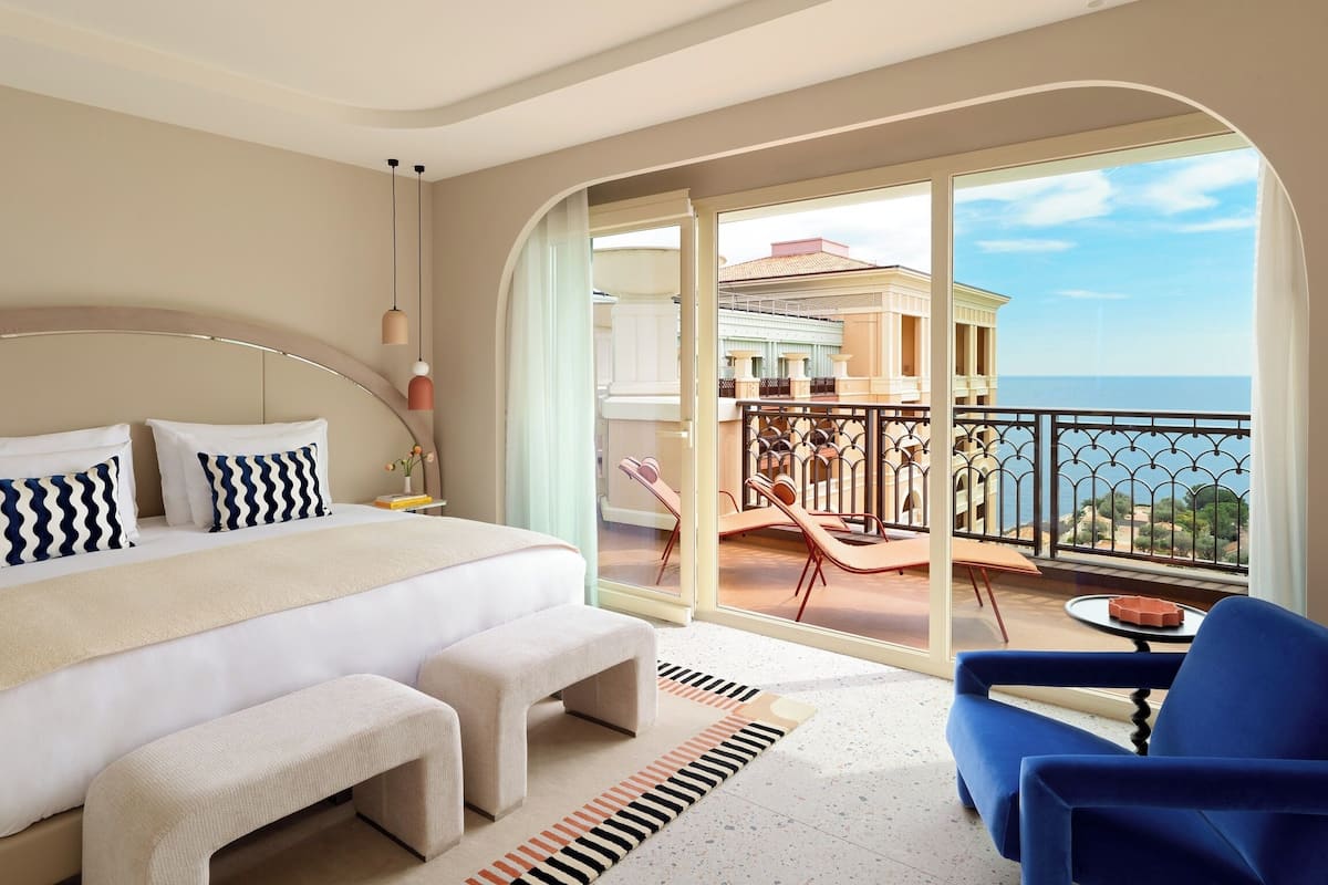Quarto com cama grande, um banco, uma cadeira azul e varanda com vista para o mar.