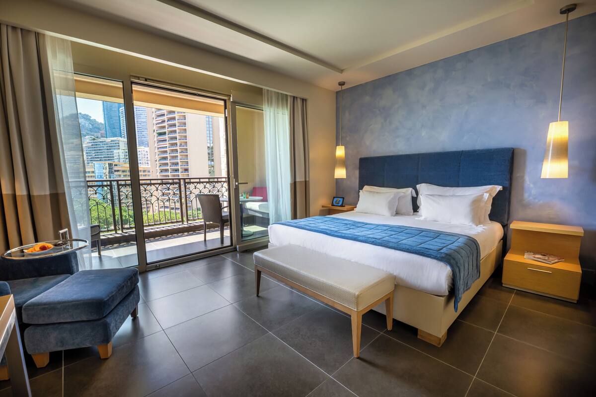 Quarto de hotel moderno com uma cama grande, um banco, uma pequena mesa e uma varanda com vista para a cidade.