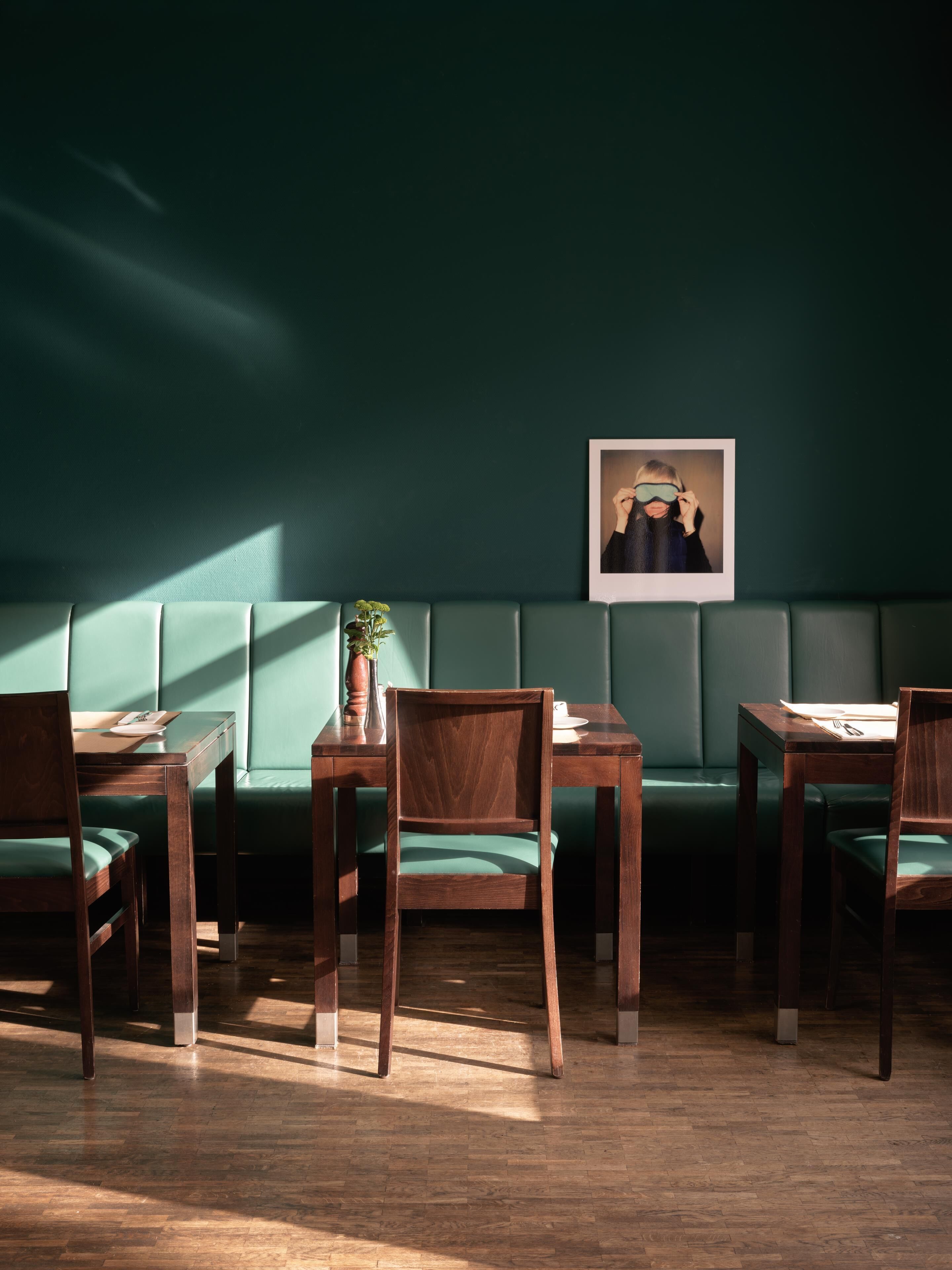 Interior de restaurante com mesas e cadeiras de madeira, um banco acolchoado verde e uma fotografia emoldurada na parede.
