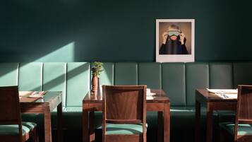 Interior de restaurante com mesas e cadeiras de madeira, um banco acolchoado verde e uma fotografia emoldurada na parede.