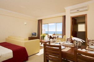 Quarto de hotel com cama, sofá, mesa de jantar posta para dois e uma grande janela com vista para o mar.