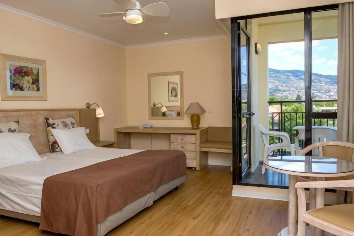 Quarto com cama, escrivaninha, cadeira e varanda com vista.