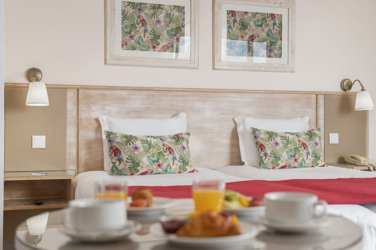 Quarto de hotel com duas camas, almofadas estampadas com flores, manta vermelha e itens de café da manhã sobre uma mesa.