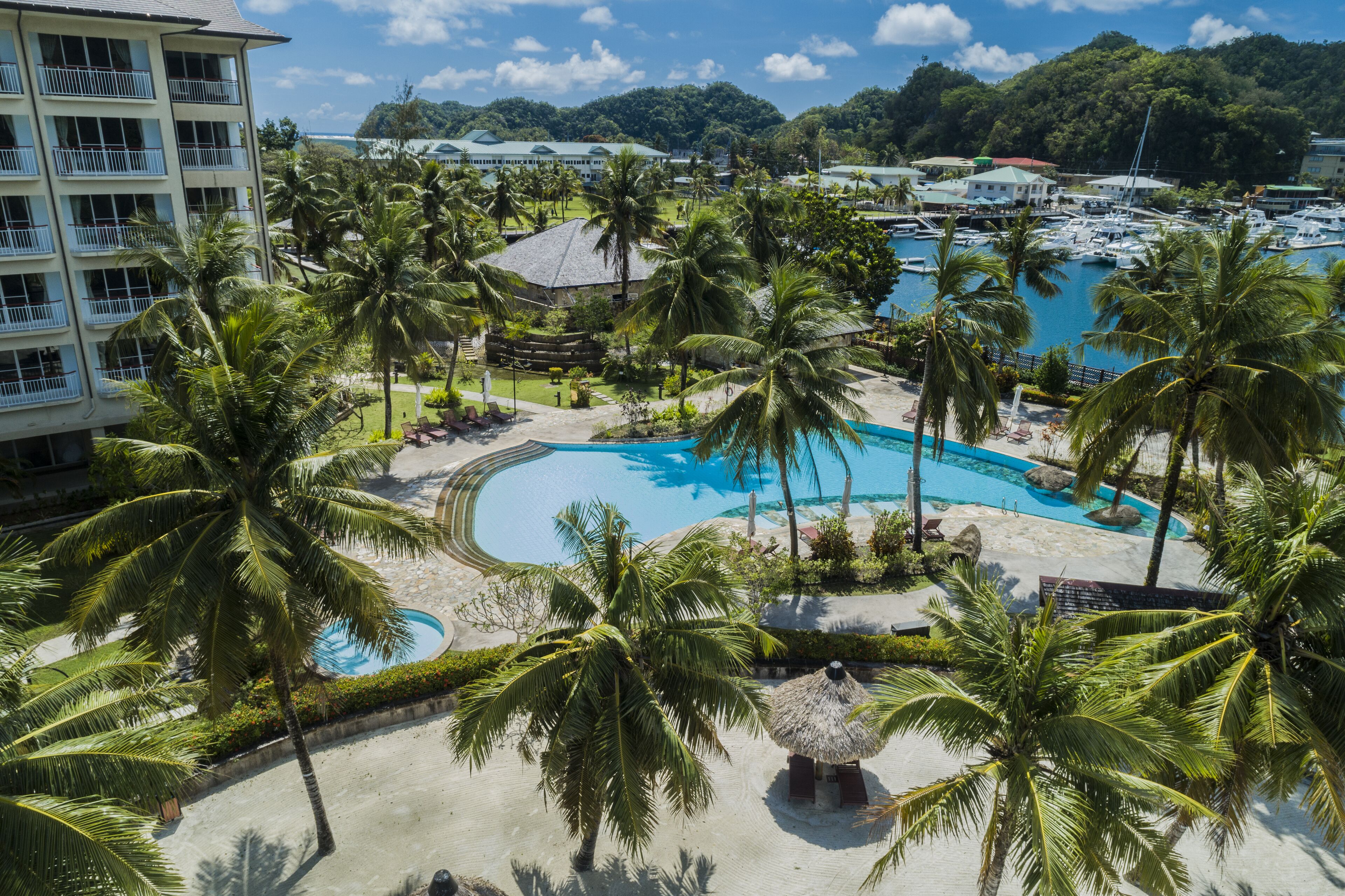Um resort com uma grande piscina cercada por palmeiras, uma marina com barcos e um edifício de hotel ao fundo.