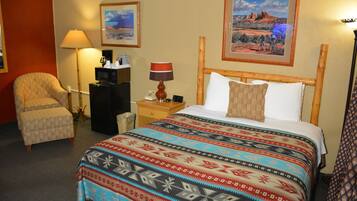 Quarto de hotel com cama, uma cadeira, escrivaninha, abajur e duas quadros na parede.