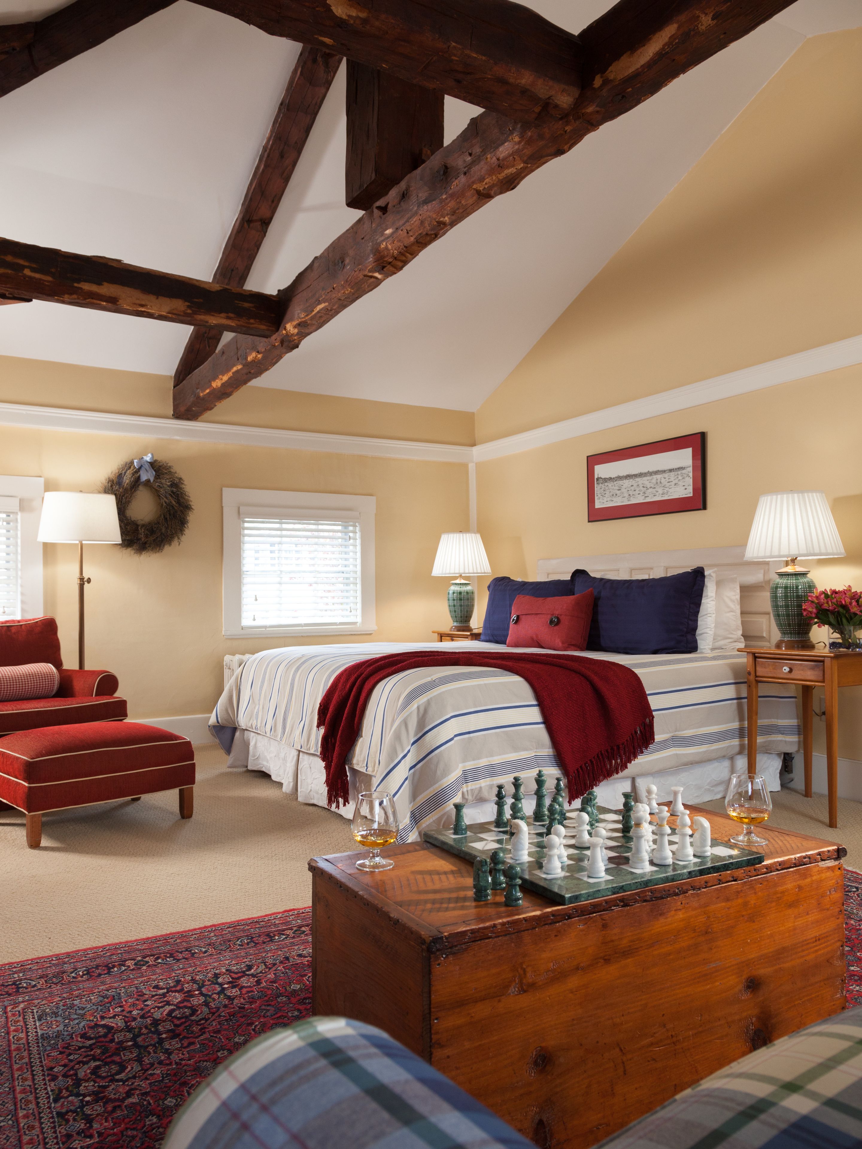 Quarto com cama, um baú de madeira, um tabuleiro de xadrez, uma poltrona vermelha, uma luminária e um quadro na parede.