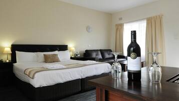 Quarto de hotel com uma cama, um sofá, uma mesa de madeira com uma garrafa de vinho e copos, e um ventilador de teto.