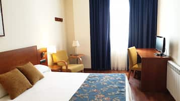 Quarto de hotel com cama, duas cadeiras, uma mesa com computador, luminária e janela com cortinas.