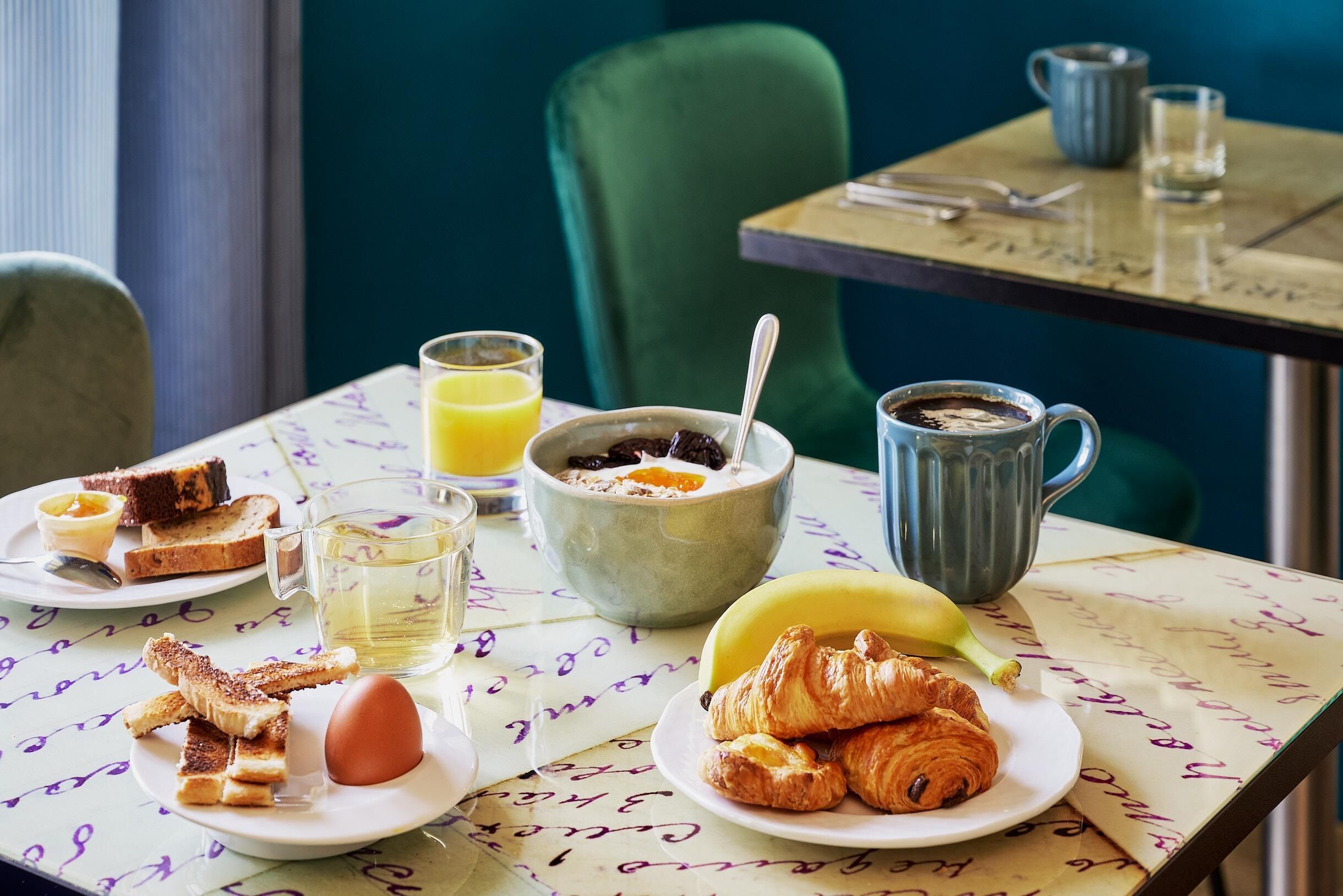 Uma mesa de café da manhã com torradas, croissants, uma banana, uma tigela de cereal, um copo de suco de laranja, uma xícara de café e uma jarra de chá.