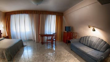 Quarto de hotel com cama, sofá, mesa de jantar com cadeiras, televisão e luminária.