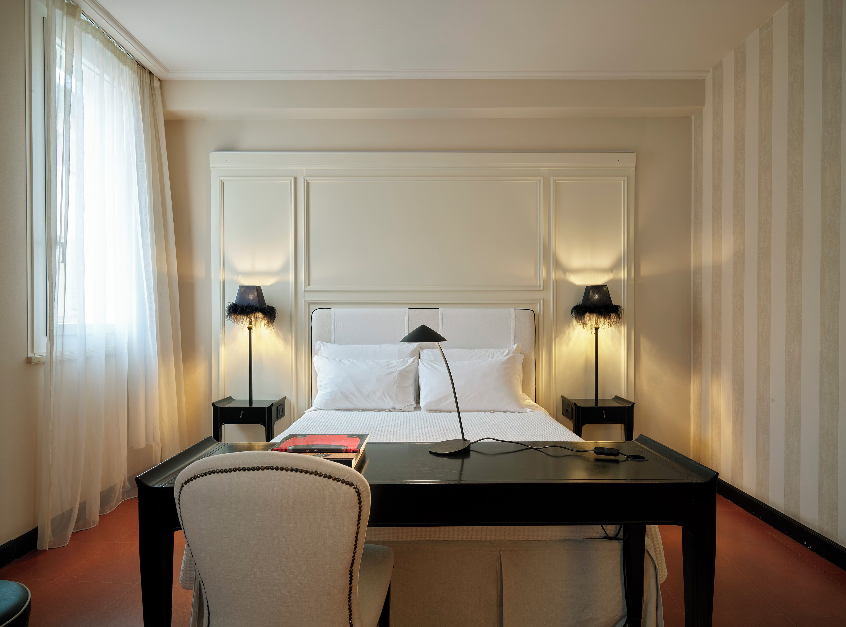 Quarto de hotel com uma cama, duas luminárias de cabeceira, uma mesa com cadeira e uma janela com cortinas leves.