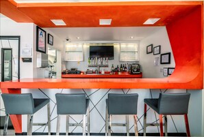 Uma área de bar moderna com balcão vermelho, quatro banquetas e uma cozinha ao fundo.