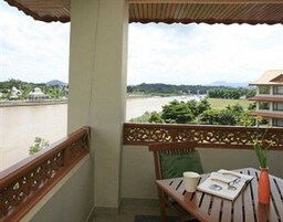 Uma varanda com vista para um rio e montanhas, uma mesa de madeira com uma xícara e um livro, e um guarda-corpo decorativo.