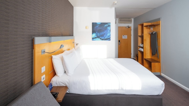 Quarto de hotel moderno com uma cama grande, cabeceira de madeira, mesa de cabeceira, guarda-roupa e uma obra de arte na parede.