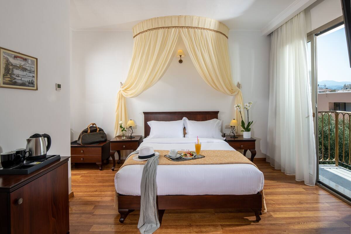 Quarto de hotel com uma cama grande, uma cômoda de madeira, uma mesa de cabeceira com abajur, uma janela com cortinas e uma varanda com vista.