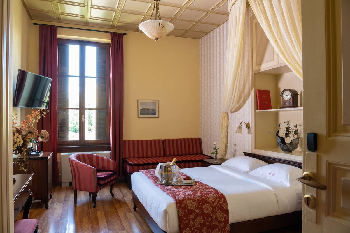 Quarto de hotel com cama, escrivaninha com televisão, cadeira, janela com cortinas e estante de livros.