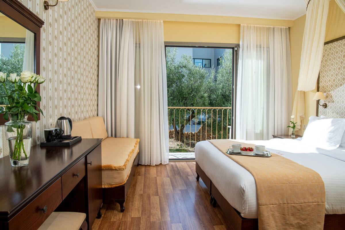 Quarto de hotel com uma cama, uma escrivaninha, uma cadeira, uma mesa pequena com um vaso de flores e vista para o exterior através de uma varanda.