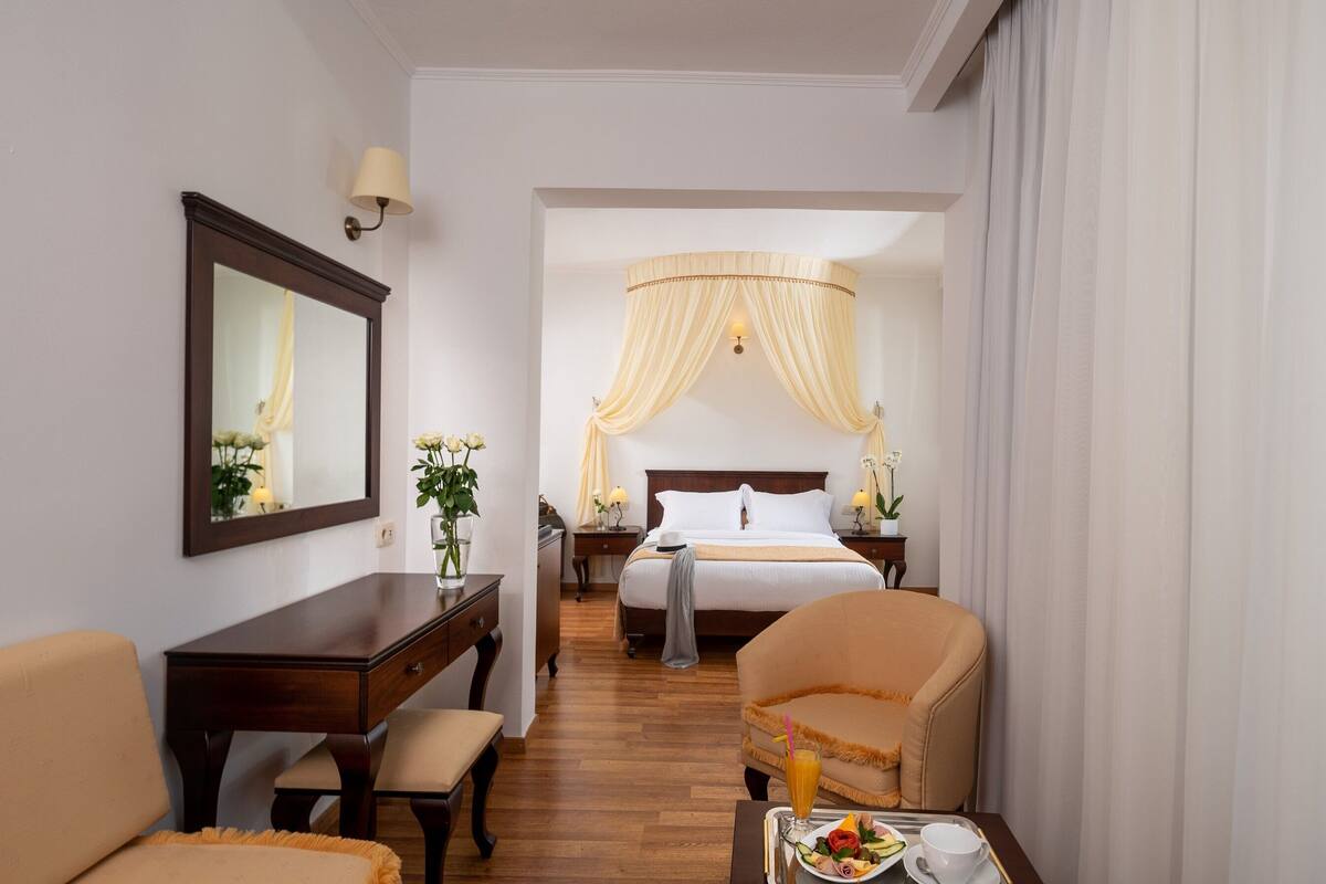 Quarto de hotel com cama, escrivaninha, cadeira, mesa de jantar e bandeja com comida, além de janela com vista para edifícios e áreas verdes.