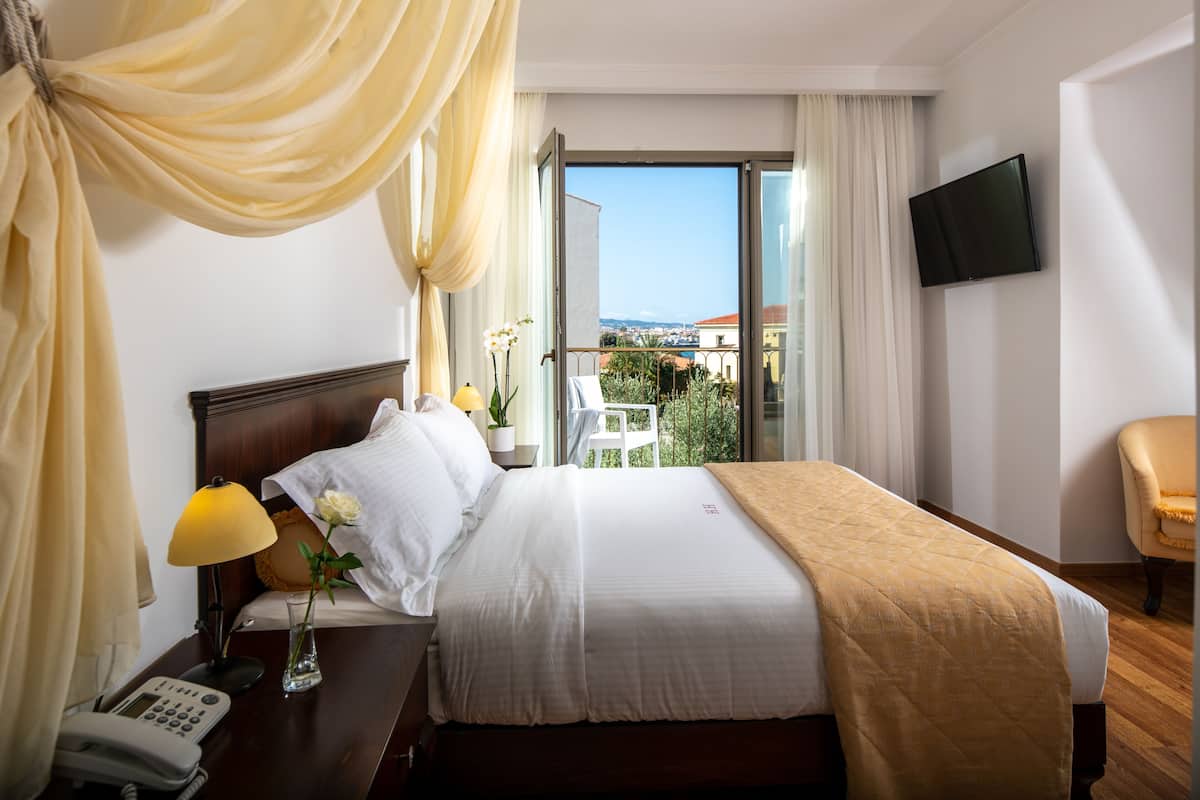 Quarto de hotel com uma cama, mesa de cabeceira com abajur e um vaso de flores, uma cadeira e uma TV fixada na parede.