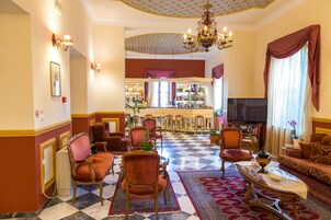 Um saguão de hotel com piso xadrez, área de estar com sofás e poltronas, um bar com adega e uma televisão.