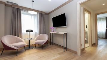 Quarto de hotel moderno com duas poltronas, uma mesa redonda pequena, televisão de tela plana na parede e uma janela grande com cortinas.