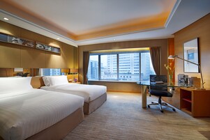 Quarto de hotel com duas camas, uma escrivaninha, uma cadeira e uma janela grande com vista para a cidade.