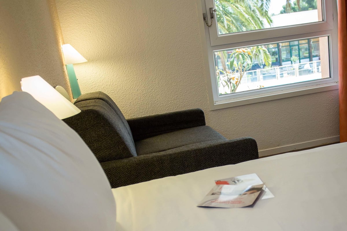 Quarto de hotel com uma cama, um pequeno sofá, uma janela com vista para palmeiras e um abajur na parede.